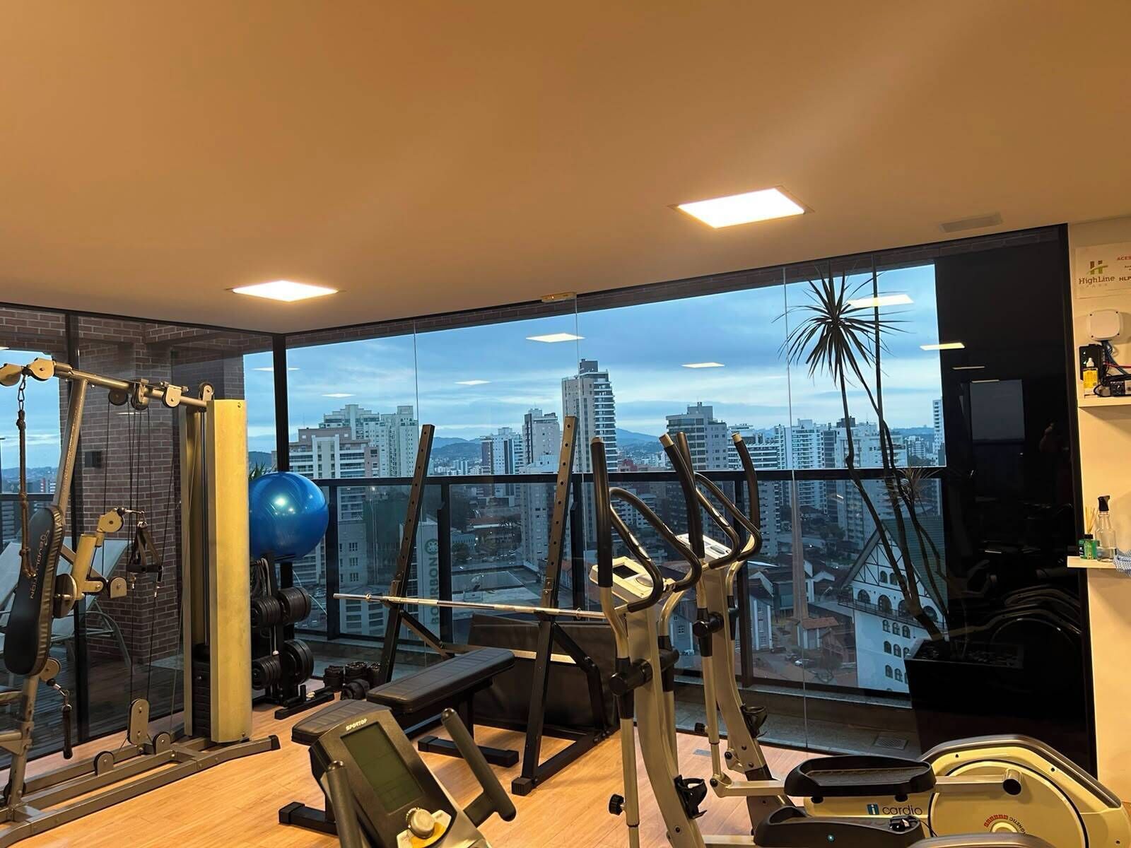 Academia com diversos equipamentos de exercício, uma grande janela com vista para a cidade e piso de madeira.