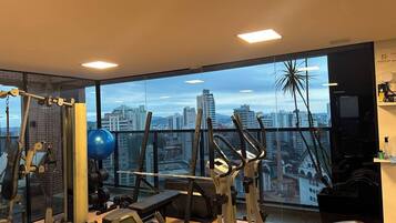 Academia com diversos equipamentos de exercício, uma grande janela com vista para a cidade e piso de madeira.
