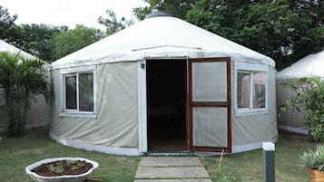 Tenda branca redonda de um único andar, com porta e janelas, cercada por grama e plantas.