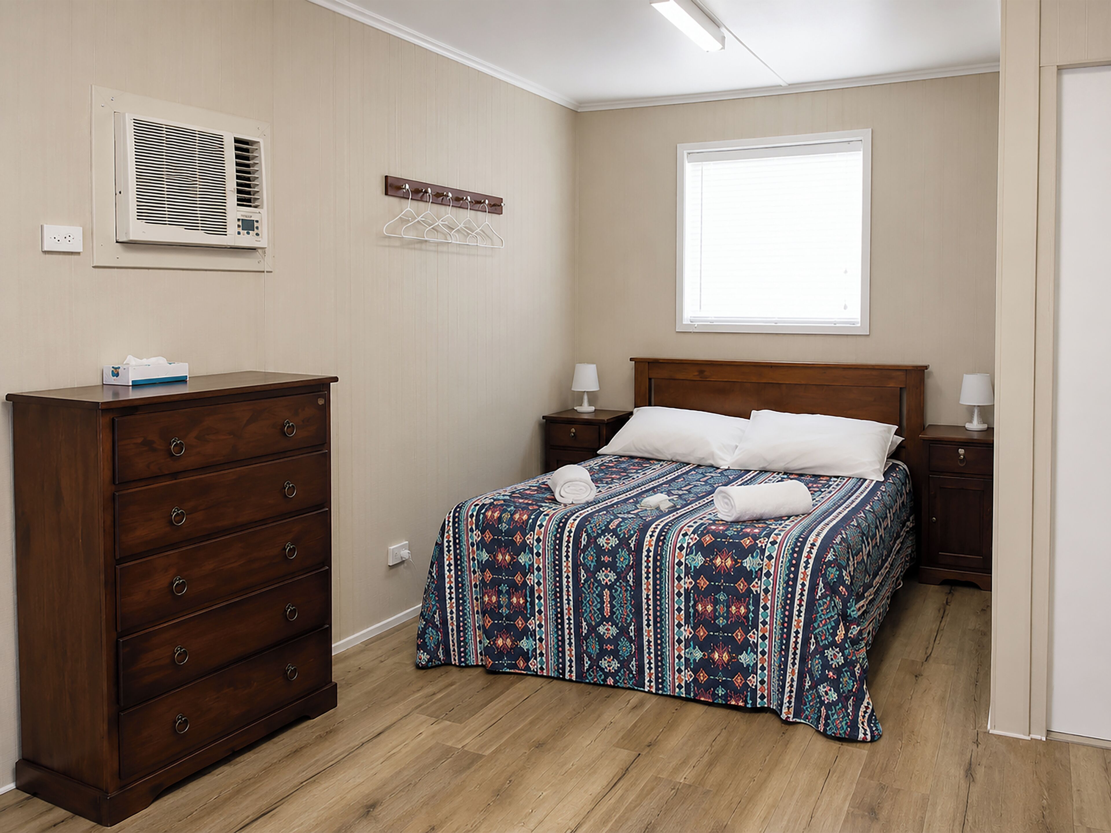 Quarto com cômoda de madeira, cama com roupa de cama estampada, janela e ar-condicionado fixado na parede.