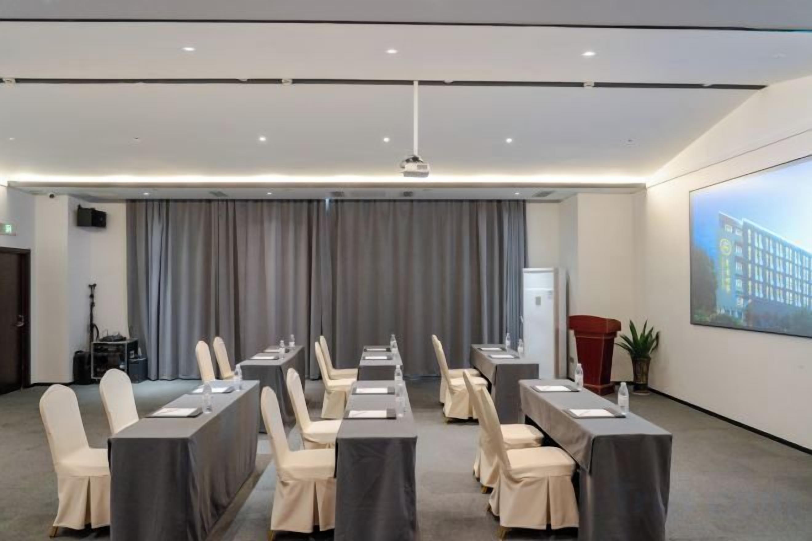 Sala de conferência com mesas e cadeiras dispostas em fileiras, um projetor e uma tela grande.