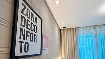 Quarto de hotel com um quadro na parede, uma cama com almofadas, uma mesa de cabeceira e uma janela com cortinas.