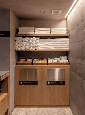 Casa de banho de hotel com um porta-toalhas, uma prateleira com artigos de higiene e um armário com sinais de 'não inflamável' e 'inflamável'.