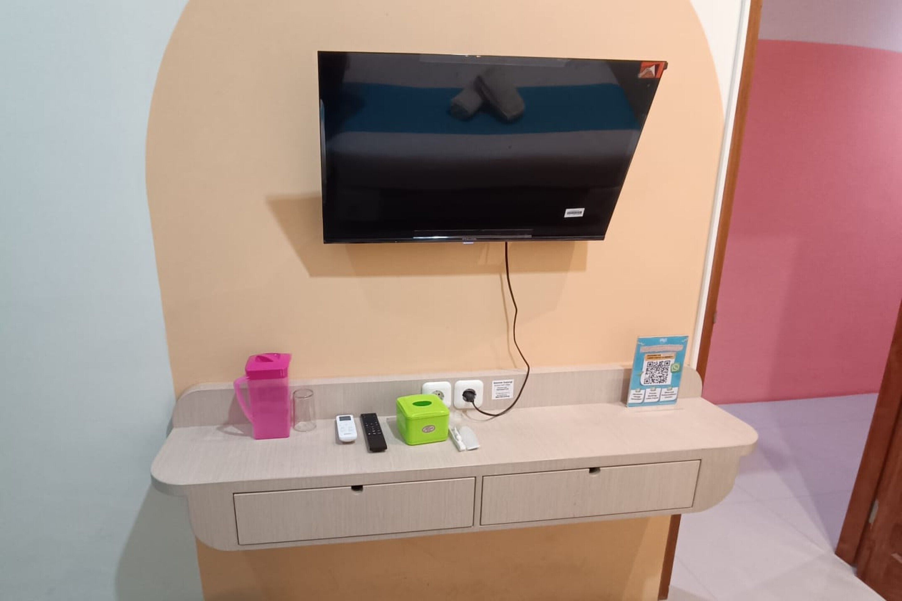 Televisão na parede acima de um balcão, com uma garrafa de água rosa, uma caixa verde, um aparelho branco e um sinal com código QR.