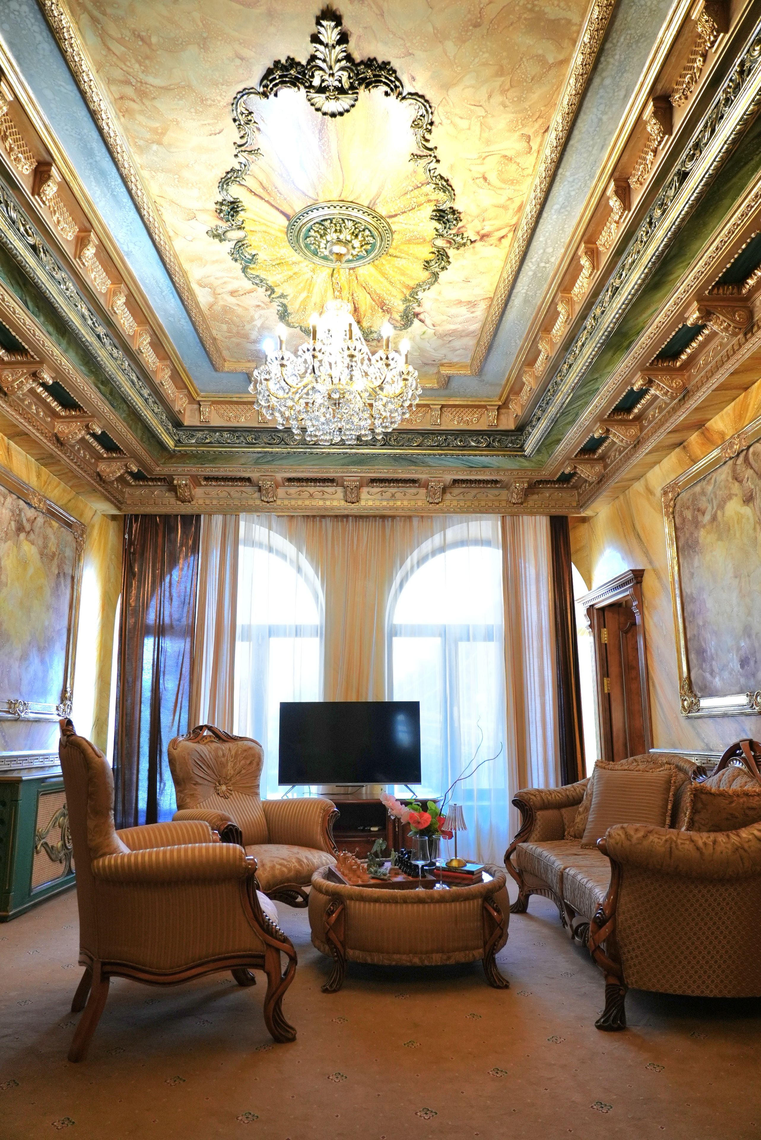 Quarto luxuoso com teto ricamente decorado, um candelabro e mobiliário clássico.