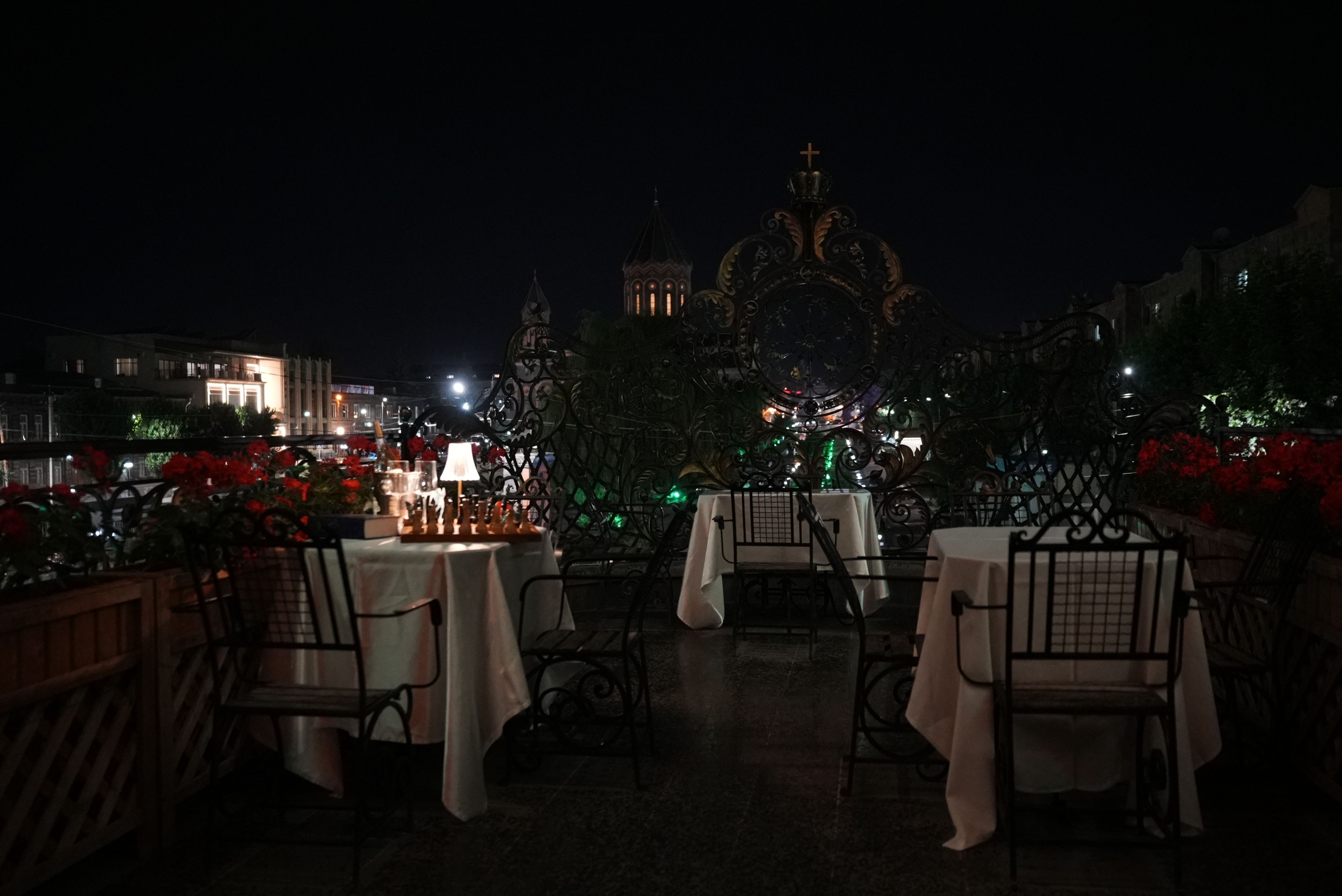 Uma área de refeições ao ar livre com mesas e cadeiras, um portão de metal decorativo e vista para um edifício com janelas iluminadas ao fundo.