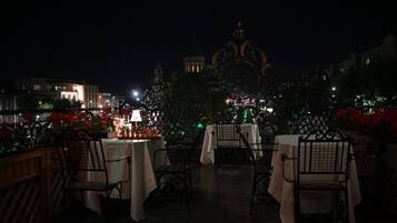 Uma área de refeições ao ar livre com mesas e cadeiras, um portão de metal decorativo e vista para um edifício com janelas iluminadas ao fundo.
