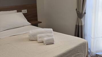 Uma cama bem arrumada com colcha estampada em florais, duas toalhas dobradas, uma cabeceira de madeira e uma mesa de cabeceira com abajur.