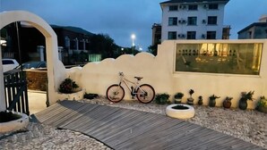 Uma entrada pavimentada com uma bicicleta, uma parede branca com uma janela e um edifício ao fundo.