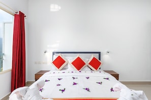 Uma cama com colcha branca, almofadas vermelhas e uma mesinha de cabeceira de madeira com luminária branca.