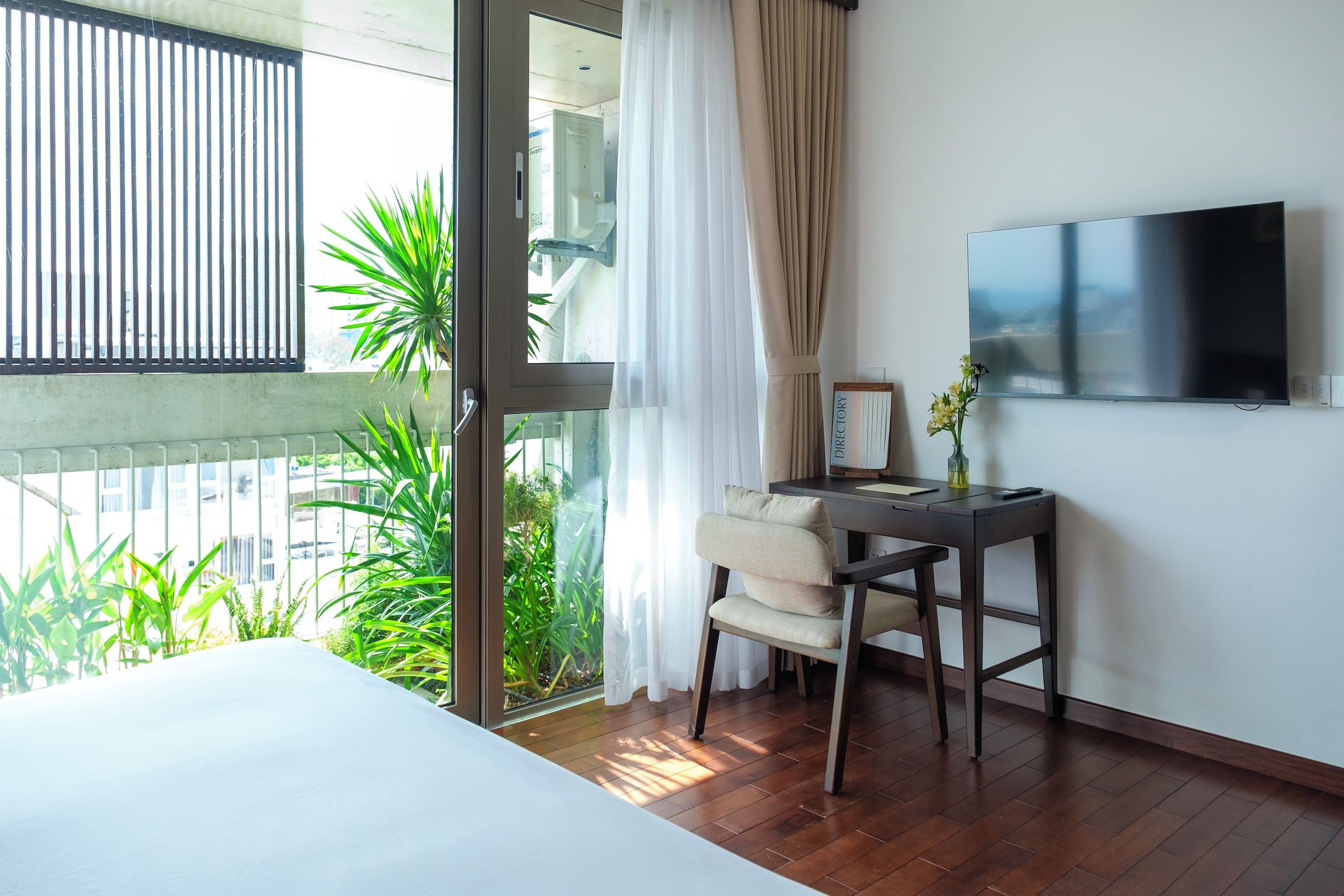 Quarto de hotel com cama, escrivaninha e cadeira, televisão na parede e vista para o verde lá fora.