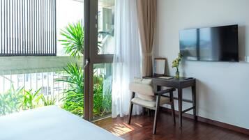 Quarto de hotel com cama, escrivaninha e cadeira, televisão na parede e vista para o verde lá fora.