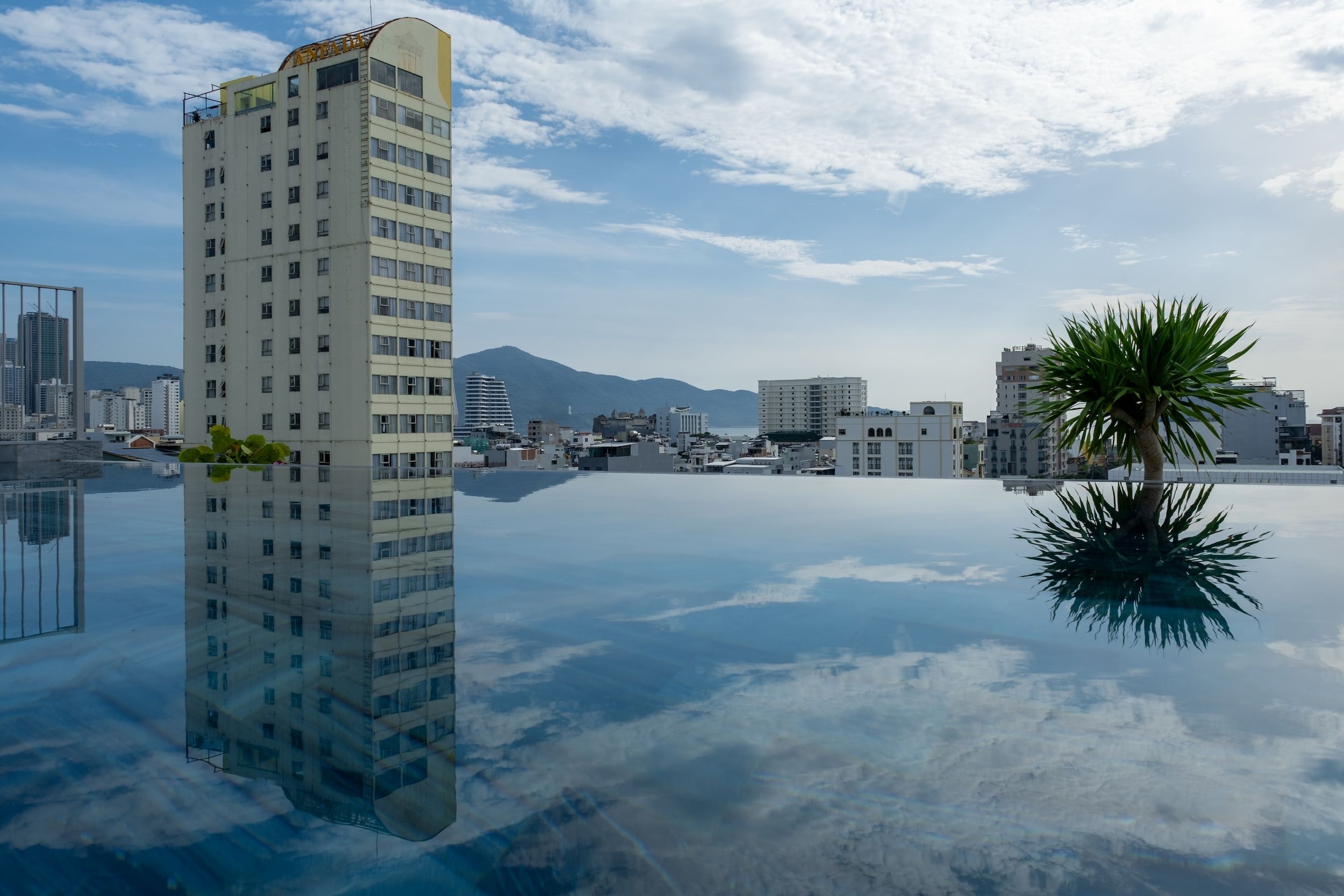 Um hotel moderno e alto refletido na água, cercado por outros edifícios e uma montanha ao fundo.
