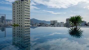 Um hotel moderno e alto refletido na água, cercado por outros edifícios e uma montanha ao fundo.