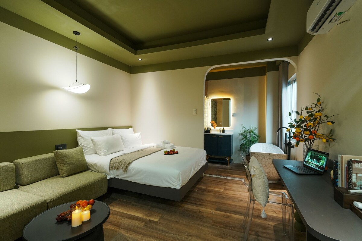 Quarto de hotel com cama, sofá, escrivaninha com um laptop, espelho e uma planta.