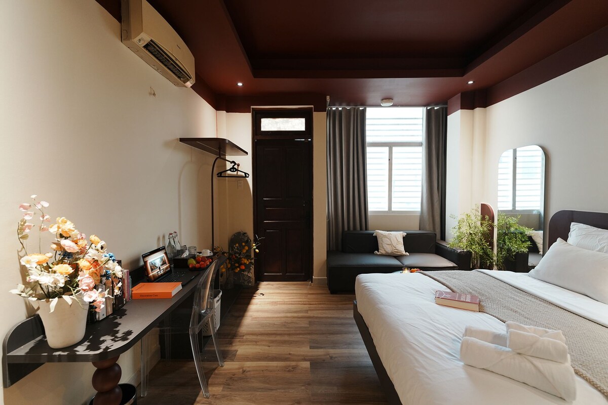 Quarto de hotel com cama, escrivaninha com computador, cadeira, abajur, janela com cortinas e porta.