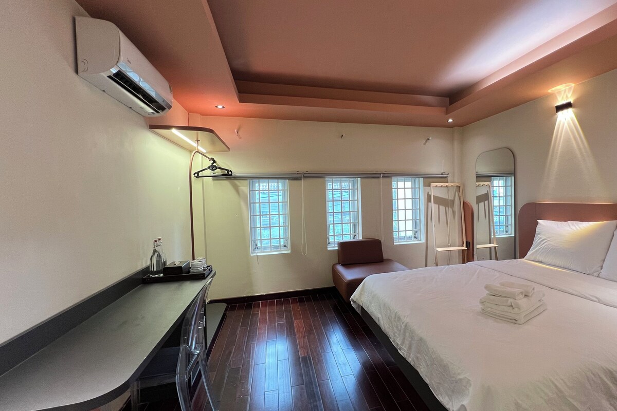Quarto de hotel com cama, mesa de cabeceira, cadeira e escrivaninha com luminária.