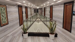 Um hall de hotel com uma mesa de vidro central, paredes de mármore, portas de madeira e plantas em vasos.