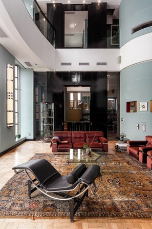 Uma sala de estar espaçosa com pé-direito alto, guarda-corpo de vidro, um sofá vermelho, uma poltrona de couro preta e um tapete estampado.