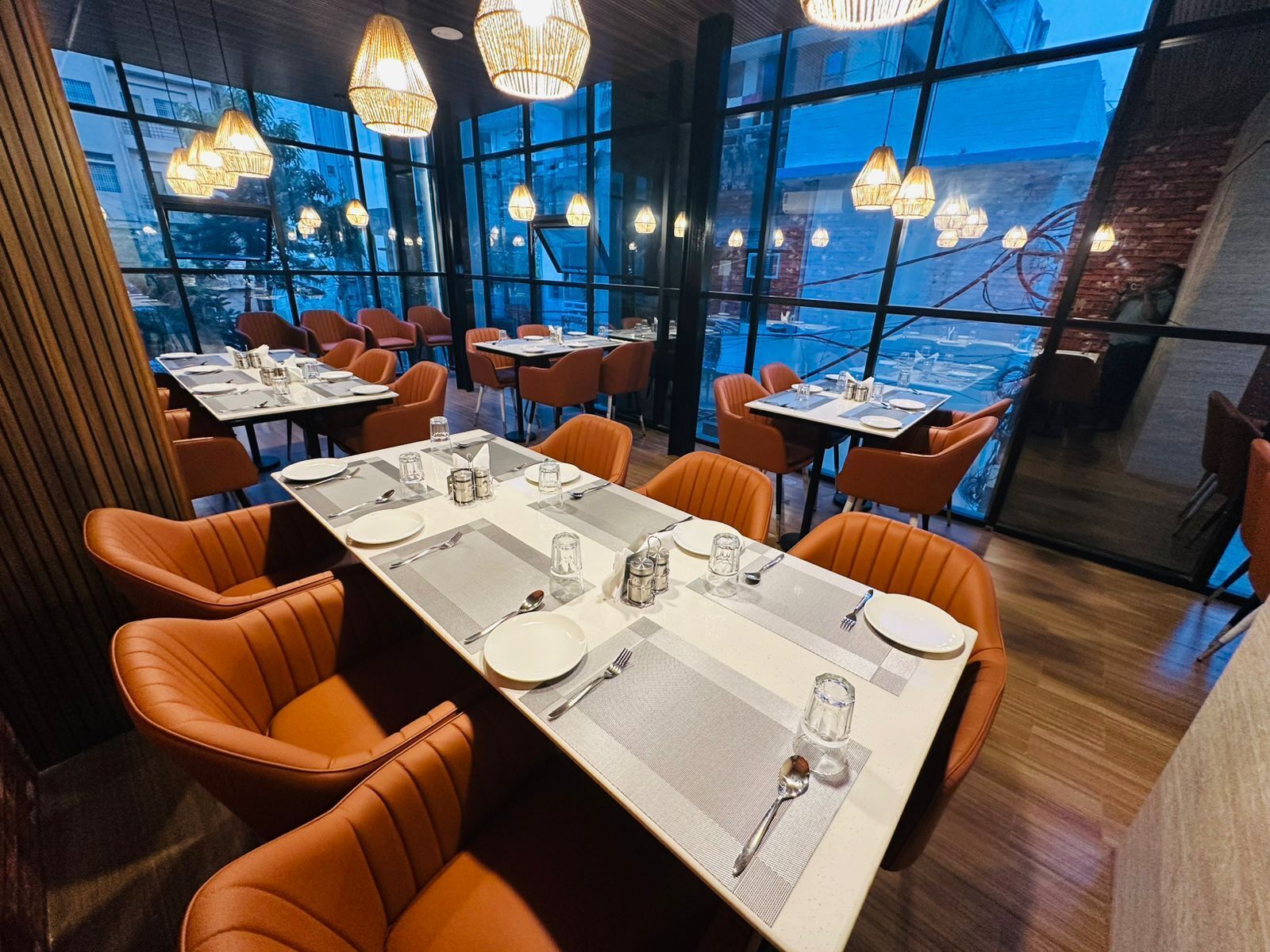 Um restaurante com mesas preparadas para refeições, grandes janelas e luminárias pendentes.