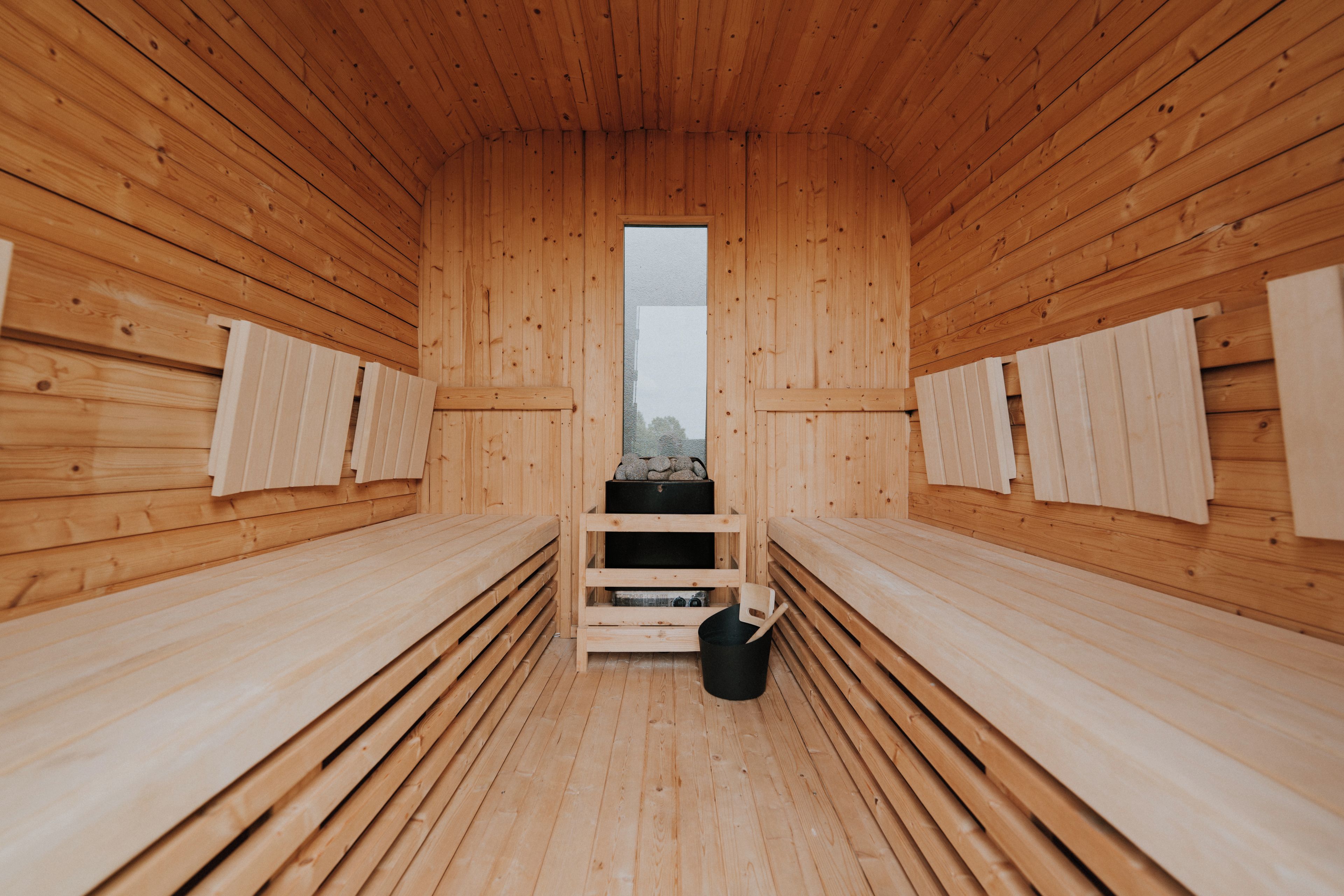 Sala de sauna com bancos de madeira e um balde preto, com vista pela janela.