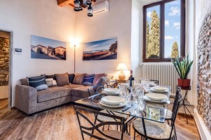 Uma sala de estar aconchegante com um sofá, uma mesa de jantar posta para quatro pessoas e duas janelas grandes com vista para árvores.