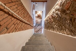 Uma escadaria com uma parede de tijolos de um lado e uma parede de pedra do outro, que leva a um quarto com uma mesa de madeira e um abajur.