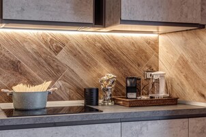 Uma bancada de cozinha com design moderno, destacando um revestimento de madeira, uma panela com espaguete cru, um recipiente cilíndrico preto e uma cafeteira de vidro.