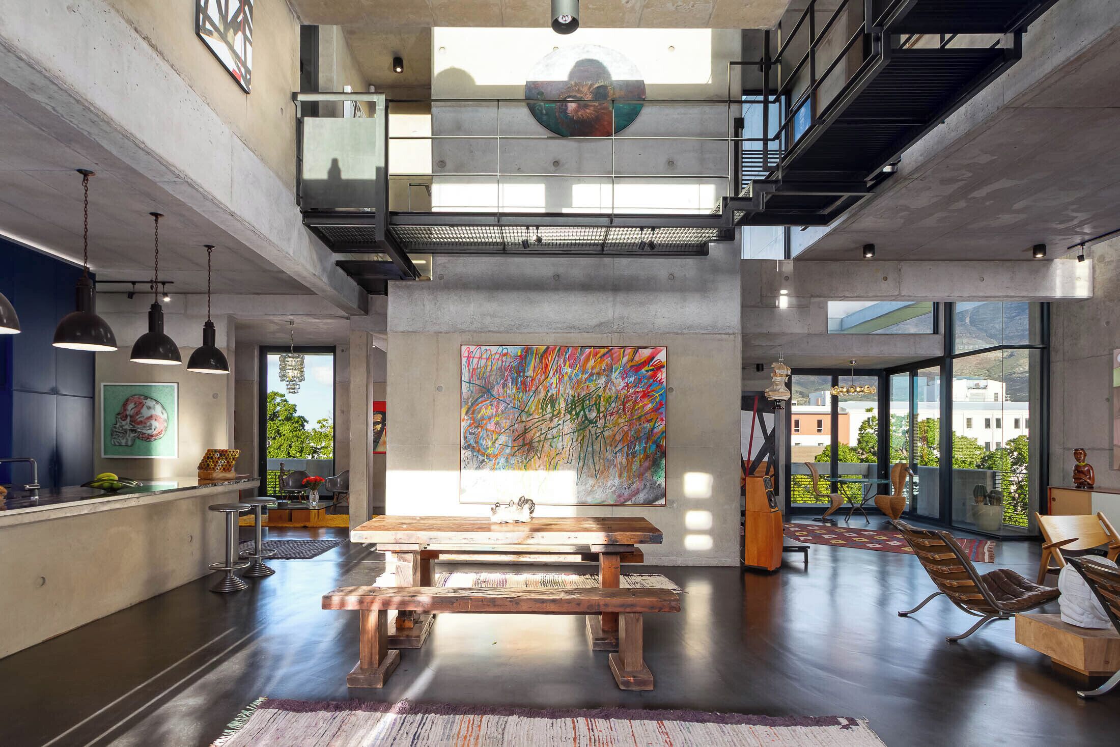 Um interior espaçoso com teto alto, uma grande pintura abstrata, uma mesa de madeira e vista para o exterior através de grandes janelas.