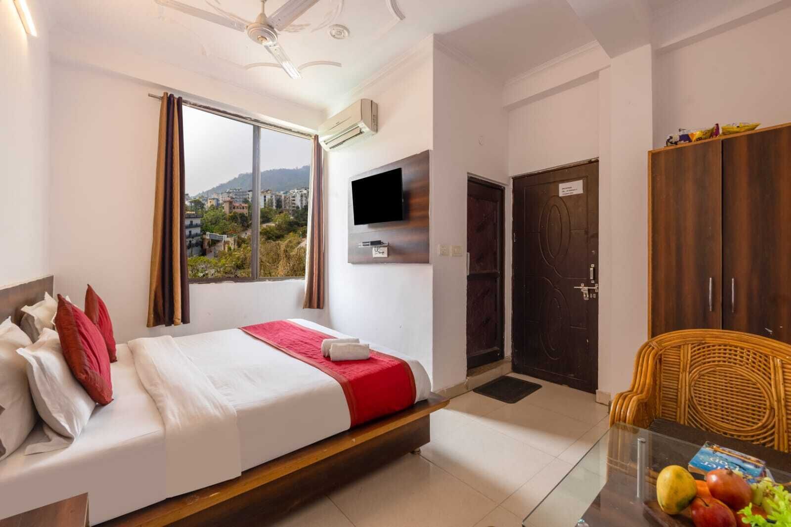 Quarto de hotel com cama, televisão, guarda-roupa, janela com vista para a cidade e uma cadeira de vime.