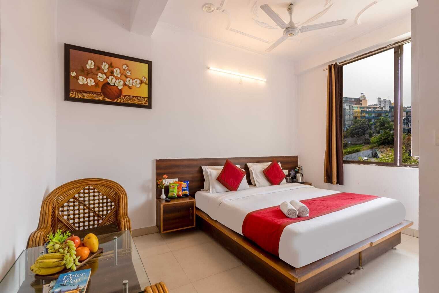 Quarto de hotel com cama, uma cadeira, uma mesa com frutas e petiscos, um quadro e vista para a cidade.