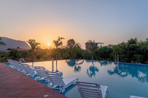 Piscina com espreguiçadeiras, cercada por vegetação e um edifício ao fundo.