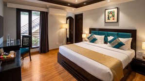 Quarto de hotel com cama, escrivaninha, cadeira, ventoinha no teto e janela com cortinas.