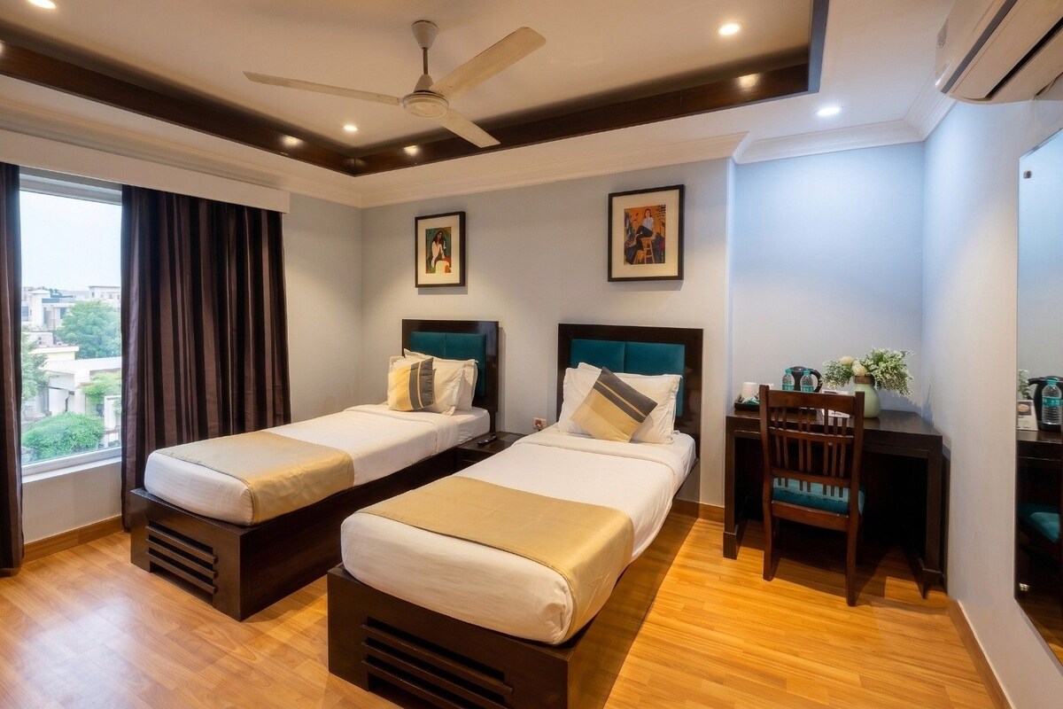 Quarto de hotel com duas camas, uma escrivaninha, uma cadeira e um ventilador de teto.