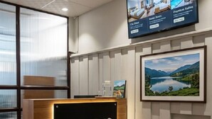 Uma área de recepção com uma mesa de madeira com a placa 'asar', um sofá cinza, uma televisão exibindo opções de quartos e uma pintura emoldurada de uma paisagem.