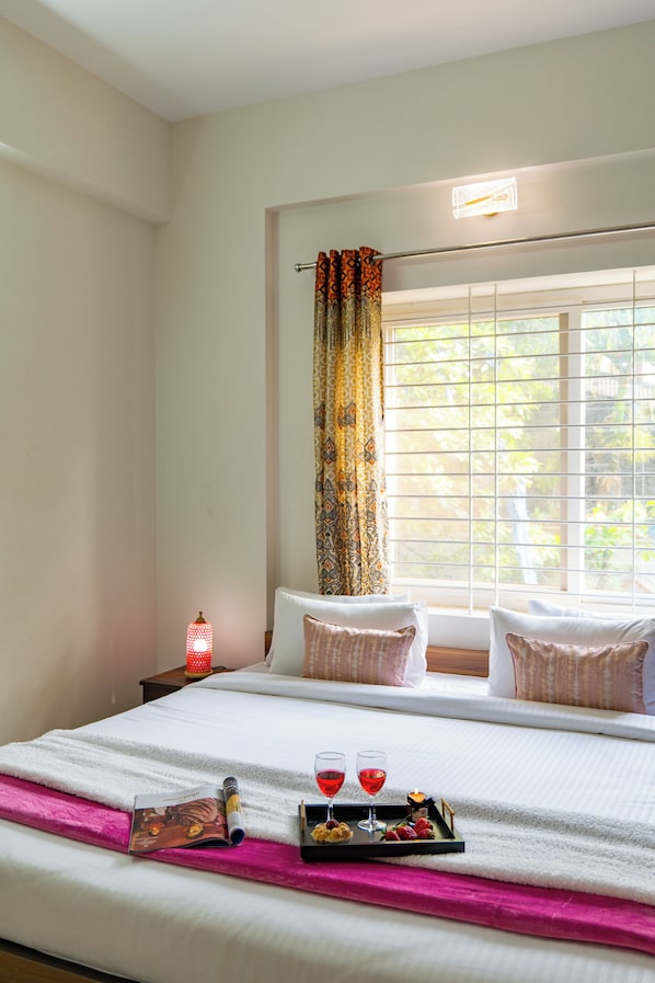 Cama bem arrumada com um tabuleiro de petiscos e bebidas, abajur e janela com cortinas.