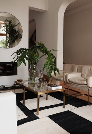 Uma sala de estar moderna com uma mesa de centro de vidro, uma planta grande em vaso, um sofá bege e um tapete preto e branco.