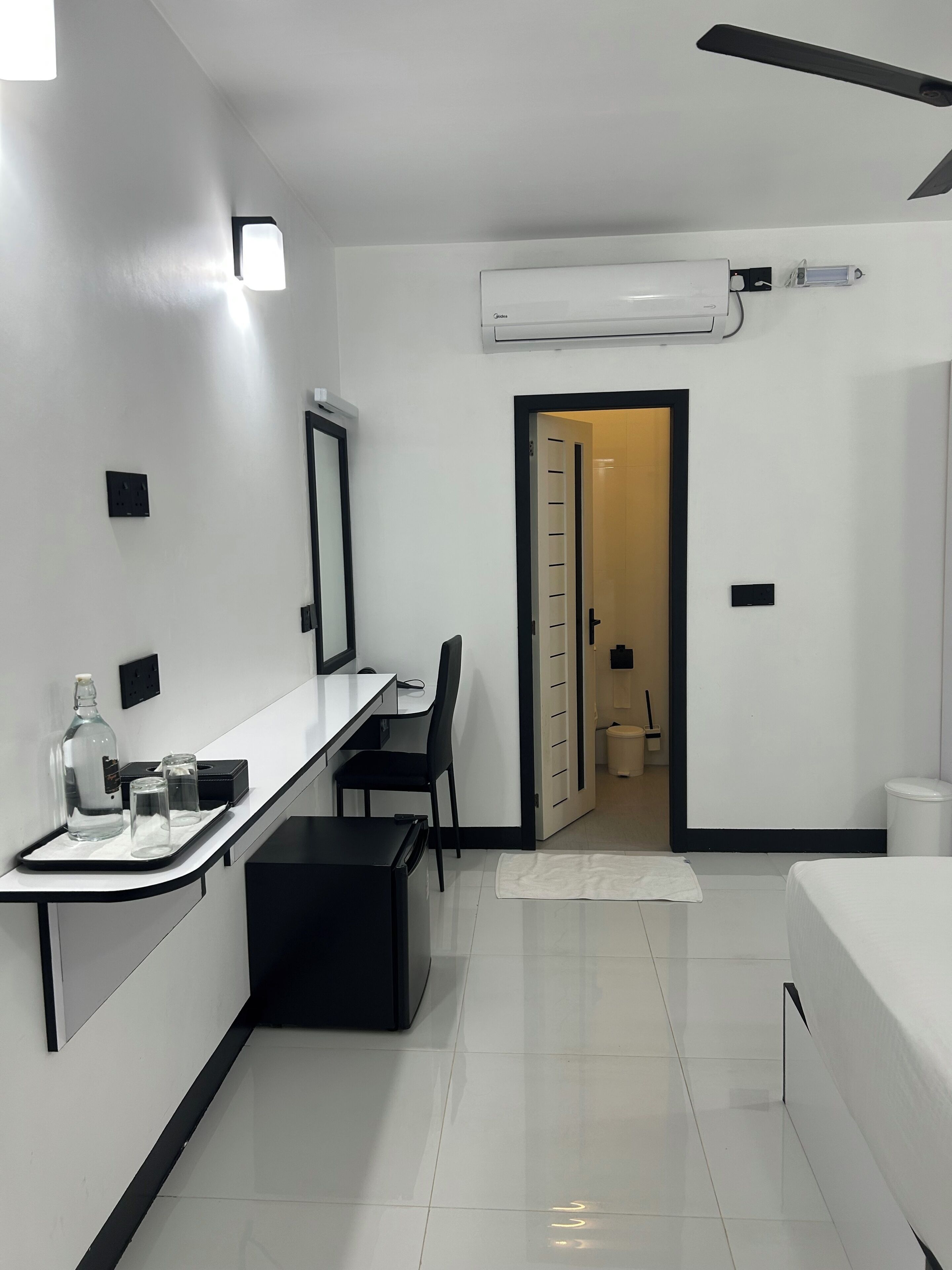 Quarto de hotel moderno com uma escrivaninha, cadeira e cama. Há um aparelho de ar condicionado fixado na parede e uma porta que dá para o banheiro.
