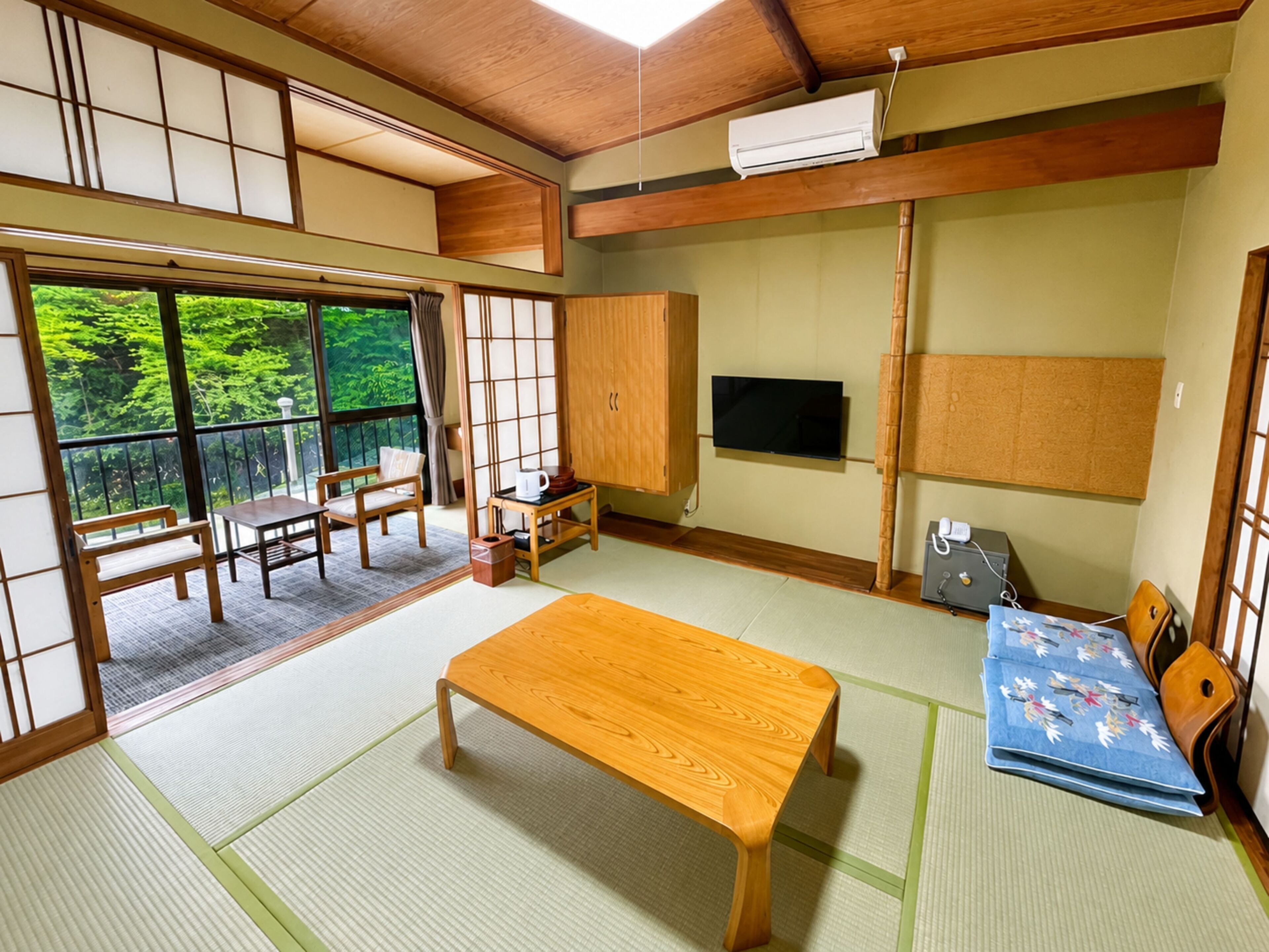 Um quarto tradicional em estilo japonês, com tatames, uma mesa de madeira baixa, televisão e vista para o verde através de grandes janelas.
