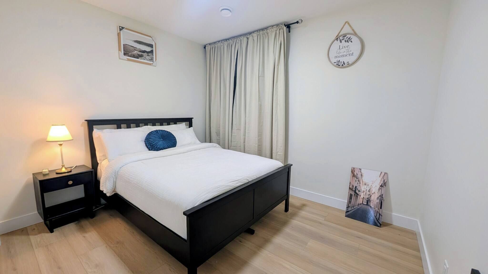 Quarto com cama, mesa de cabeceira com abajur, janela com cortinas e dois objetos decorativos na parede.