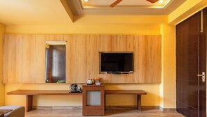 Quarto de hotel com um banco de madeira, uma mesa pequena com uma chaleira, televisão fixada na parede e uma porta de madeira grande.