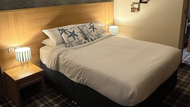 Cama bem arrumada com cabeceira, abajur e quadro na parede.