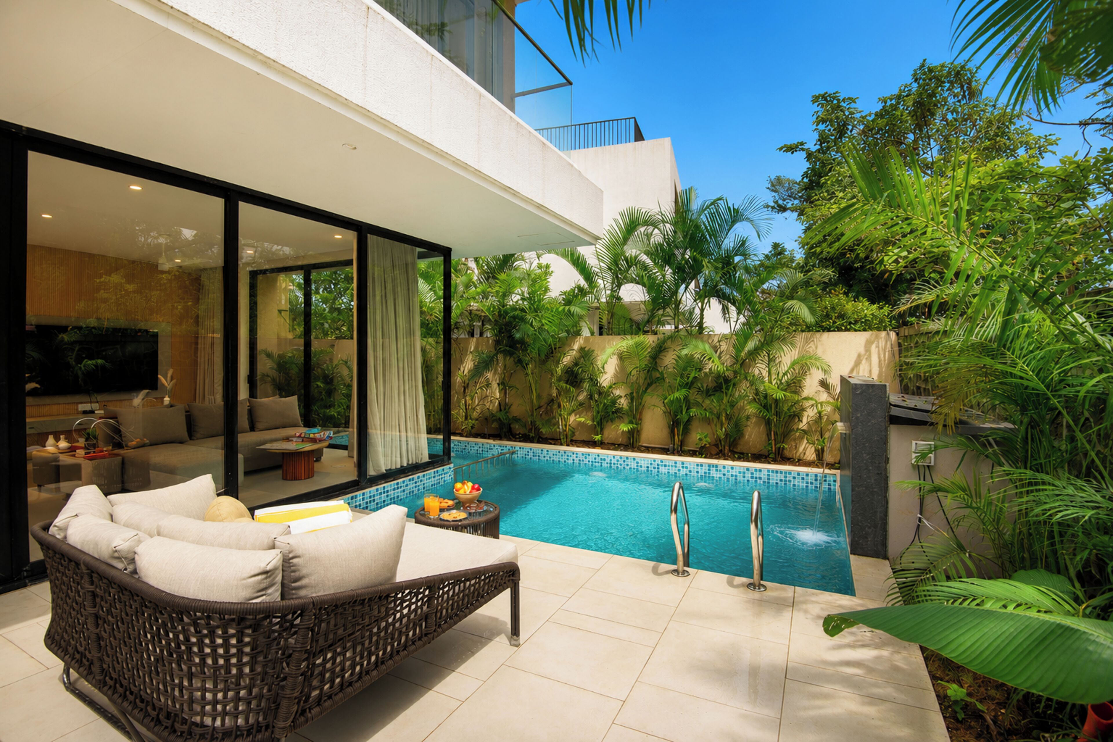 Uma área de piscina moderna ao ar livre, com um lounge envidraçado, um sofá de vime e uma mesa de jantar com frutas e bebidas.