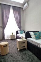 Quarto com cama, mesa de cabeceira, um banquinho pequeno e janela com cortinas.