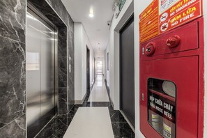 Corredor com piso de mármore, um carretel de mangueira de incêndio vermelho e uma placa de saída visível.
