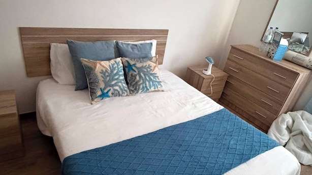 Uma cama bem arrumada com colcha azul, dois travesseiros com tema náutico, cabeceira de madeira, mesa de cabeceira com abajur e cômoda de madeira com produtos de higiene pessoal.