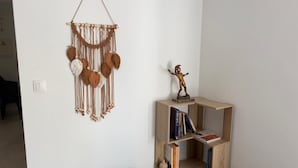 Um quarto com um painel de macramê na parede, uma estante de madeira com livros e dois pufes de couro.