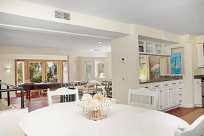 Uma sala de jantar espaçosa com mesa e cadeiras brancas, uma cozinha com armários brancos e vista para o exterior através de grandes janelas.
