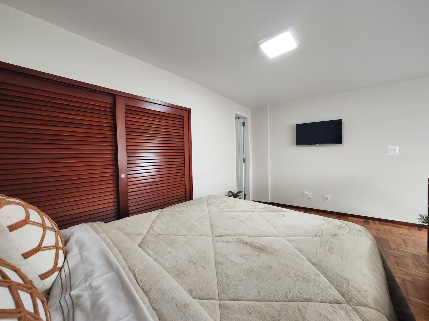 Um quarto com uma cama, um guarda-roupa de madeira, uma televisão na parede e uma porta que dá para outro cômodo.