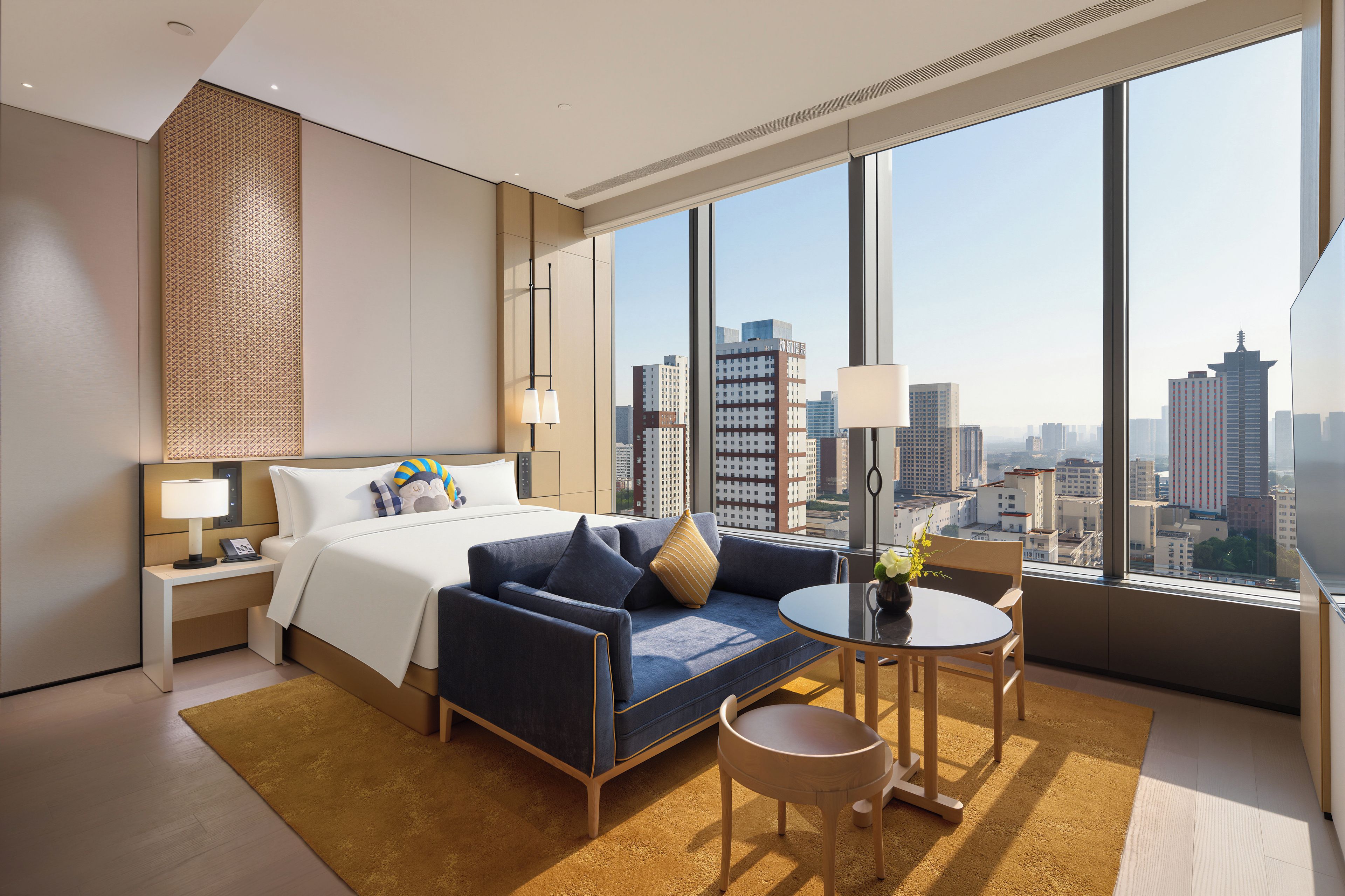 Quarto de hotel moderno com cama grande, sofá, mesinha e vista para a paisagem urbana.